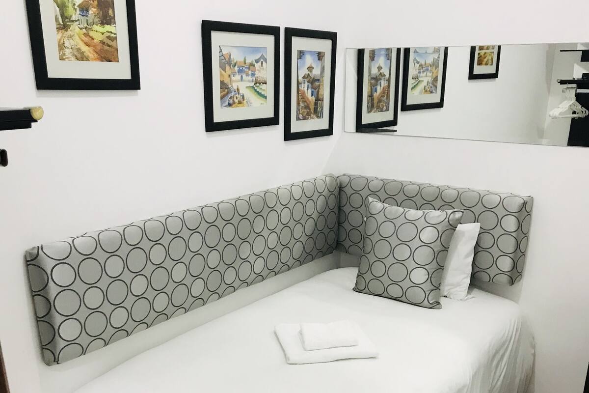 Uma cama bem arrumada com edredom branco e almofadas, cabeceira com padrão geométrico e várias obras de arte emolduradas na parede.