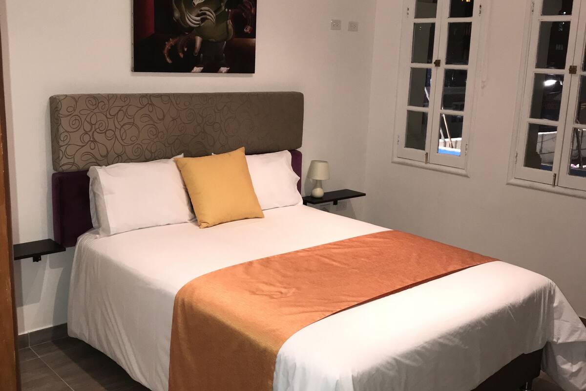 Uma cama bem arrumada com colcha branca e laranja, cabeceira com design estampado e um quadro na parede acima da cama.
