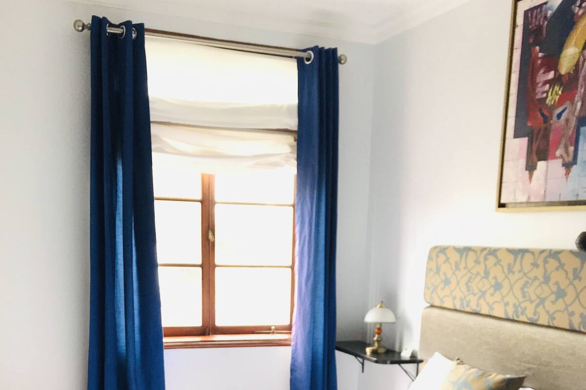 Cama bem arrumada com lençóis brancos, mesa de cabeceira com abajur e janela com cortinas azuis.