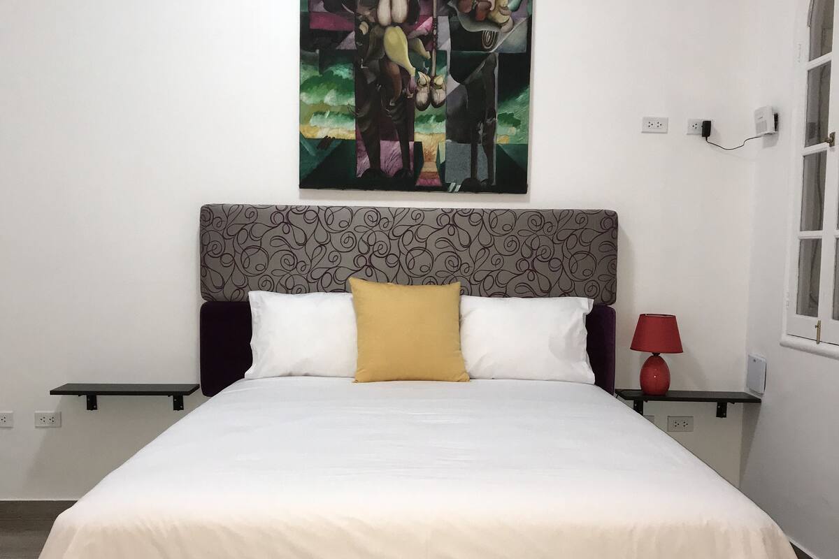 Quarto com uma cama grande, cabeceira com padrão decorativo, mesa de cabeceira com abajur vermelho e um quadro na parede.