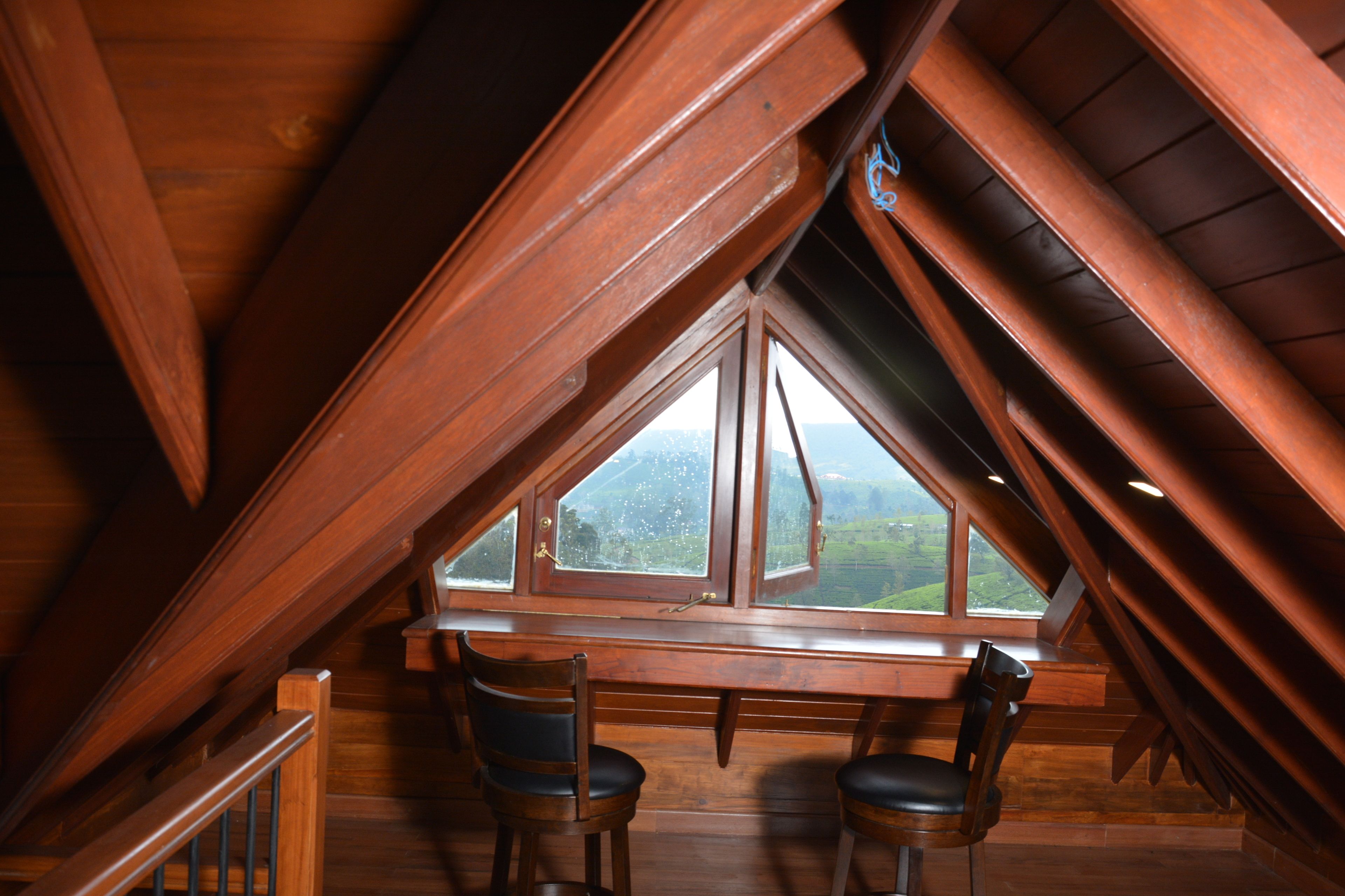 Quarto de sótão com chão de madeira, teto inclinado, duas cadeiras de madeira e uma grande janela com vista para uma paisagem verde.