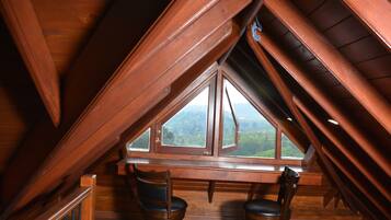 Quarto de sótão com chão de madeira, teto inclinado, duas cadeiras de madeira e uma grande janela com vista para uma paisagem verde.