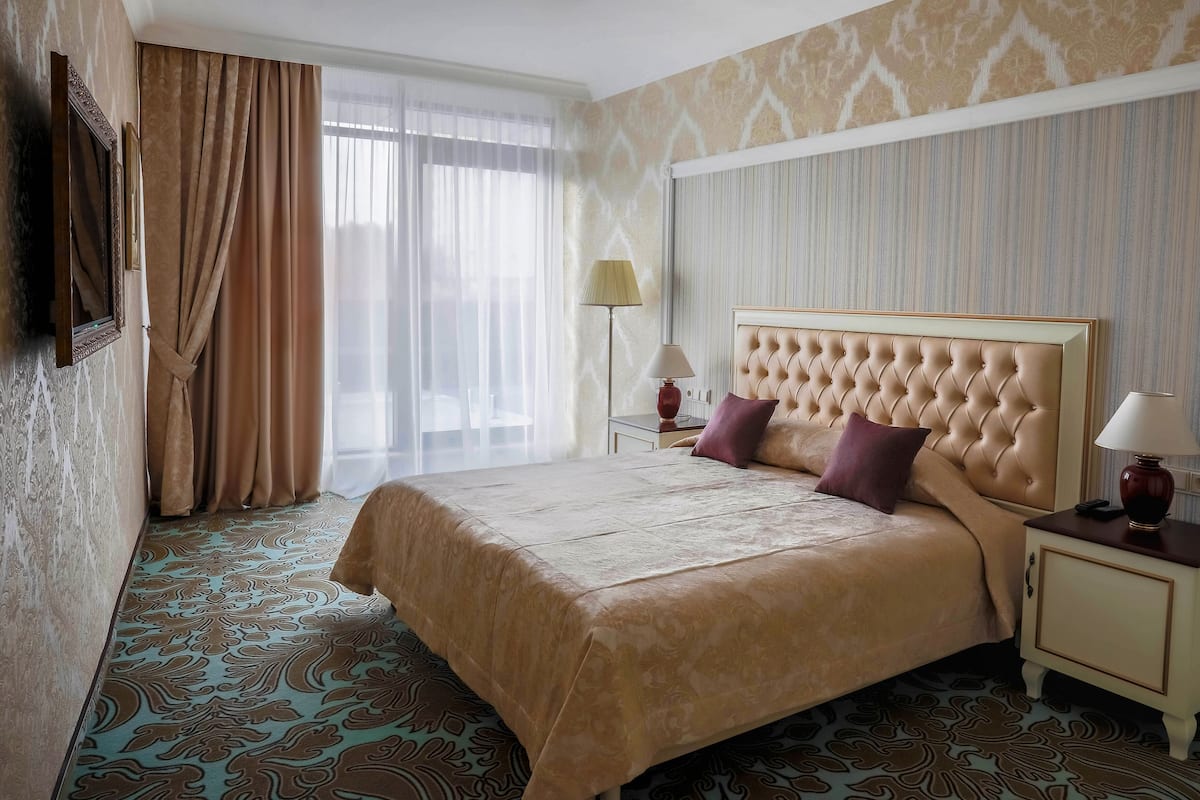 Quarto de hotel com uma cama grande, duas mesas de cabeceira com abajures, uma TV fixada na parede e um tapete estampado.