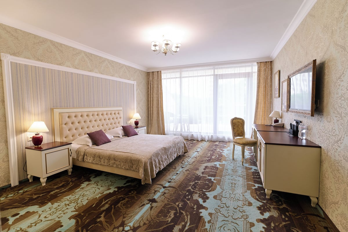 Quarto de hotel com uma cama grande, uma escrivaninha, uma cadeira, uma mesa de cabeceira com abajur e uma janela ampla com cortinas.