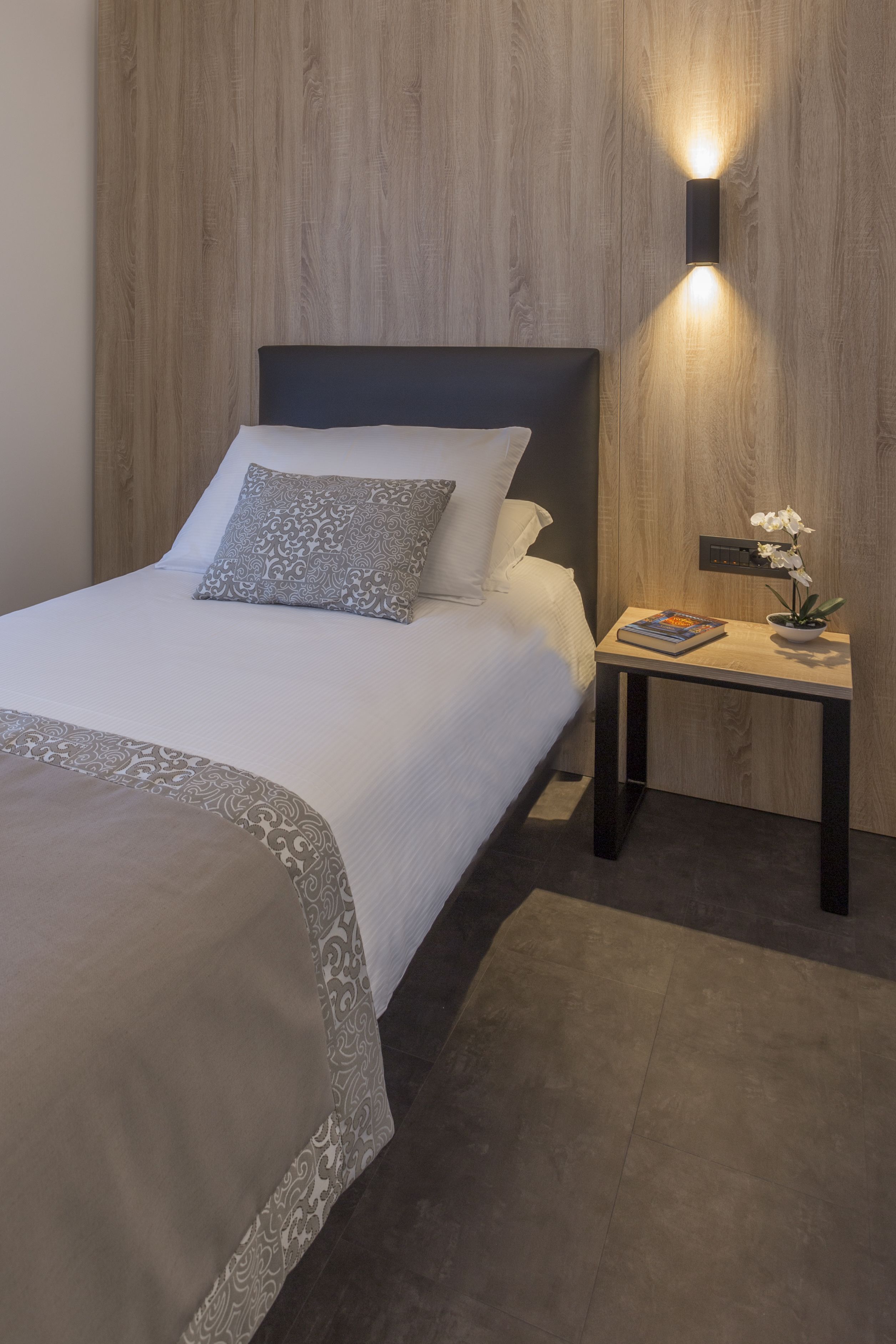 Cama bem arrumada com colcha estampada e travesseiros, mesa de cabeceira com um livro e um vaso de flores, e luminária fixada na parede.