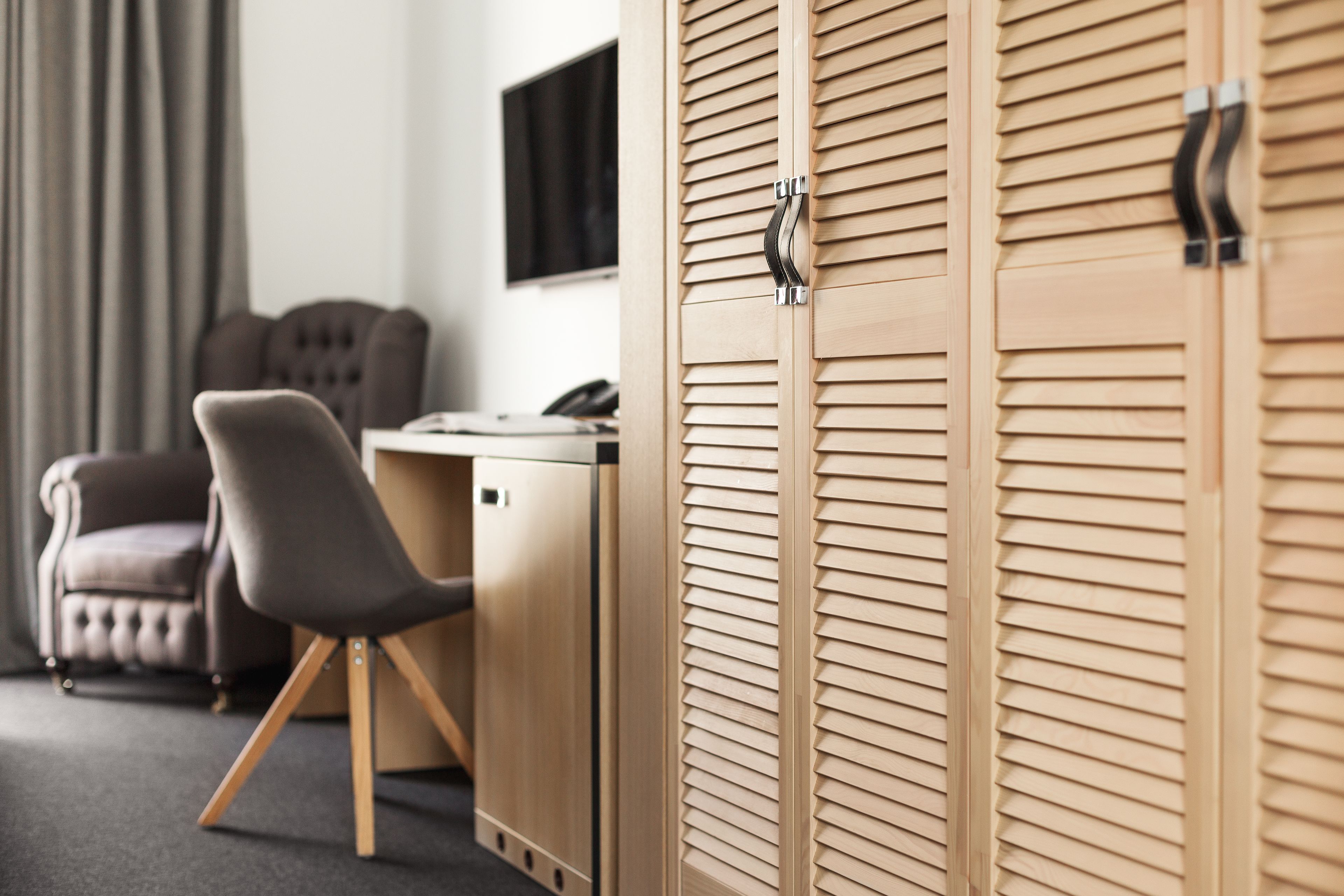 Quarto de hotel com escrivaninha, cadeira, televisão e guarda-roupa com portas venezianas.