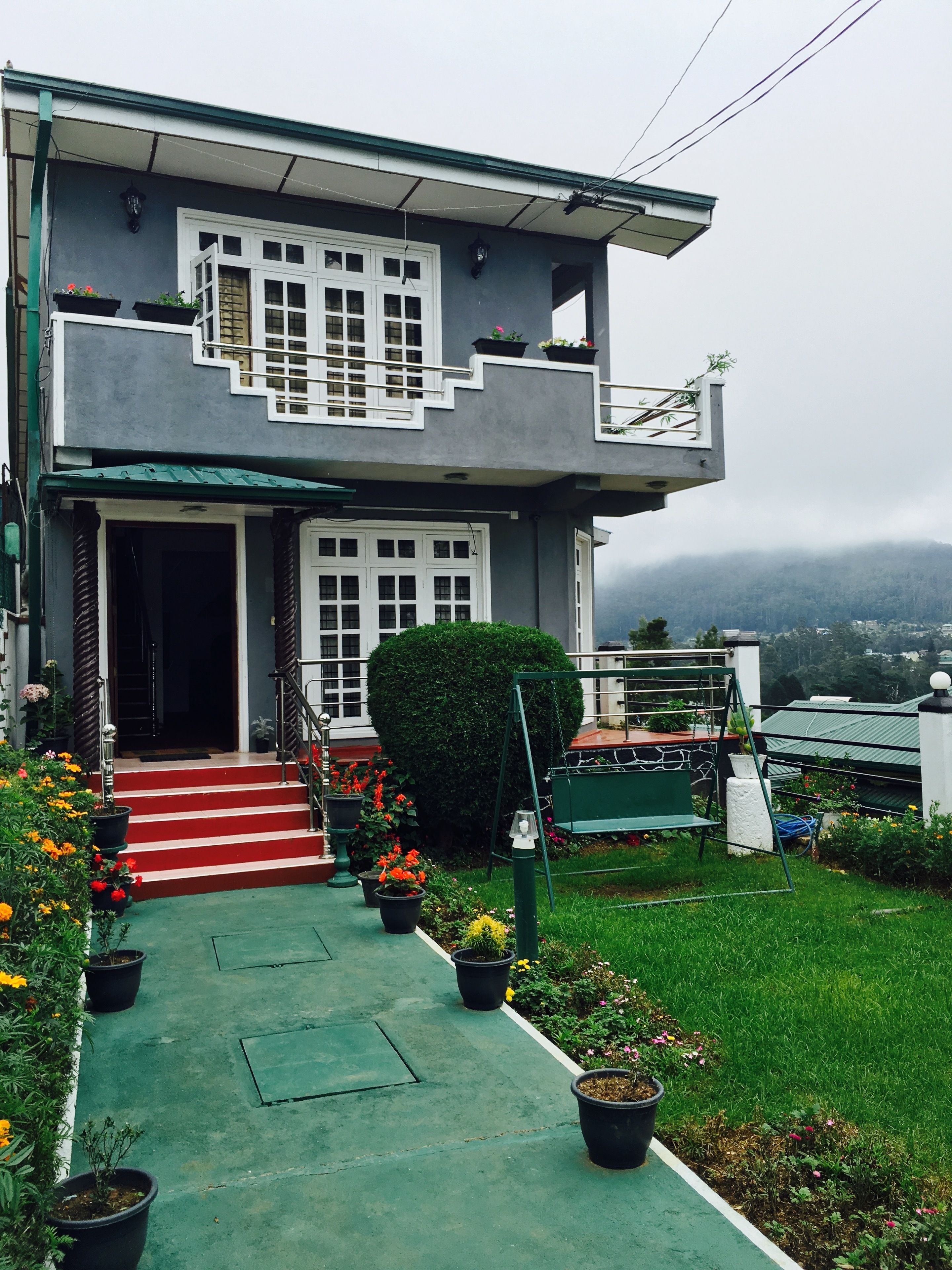 Uma casa de dois andares com telhado verde, paredes externas brancas e escadaria vermelha e branca que leva à entrada. Há plantas em vasos e flores no jardim frontal.