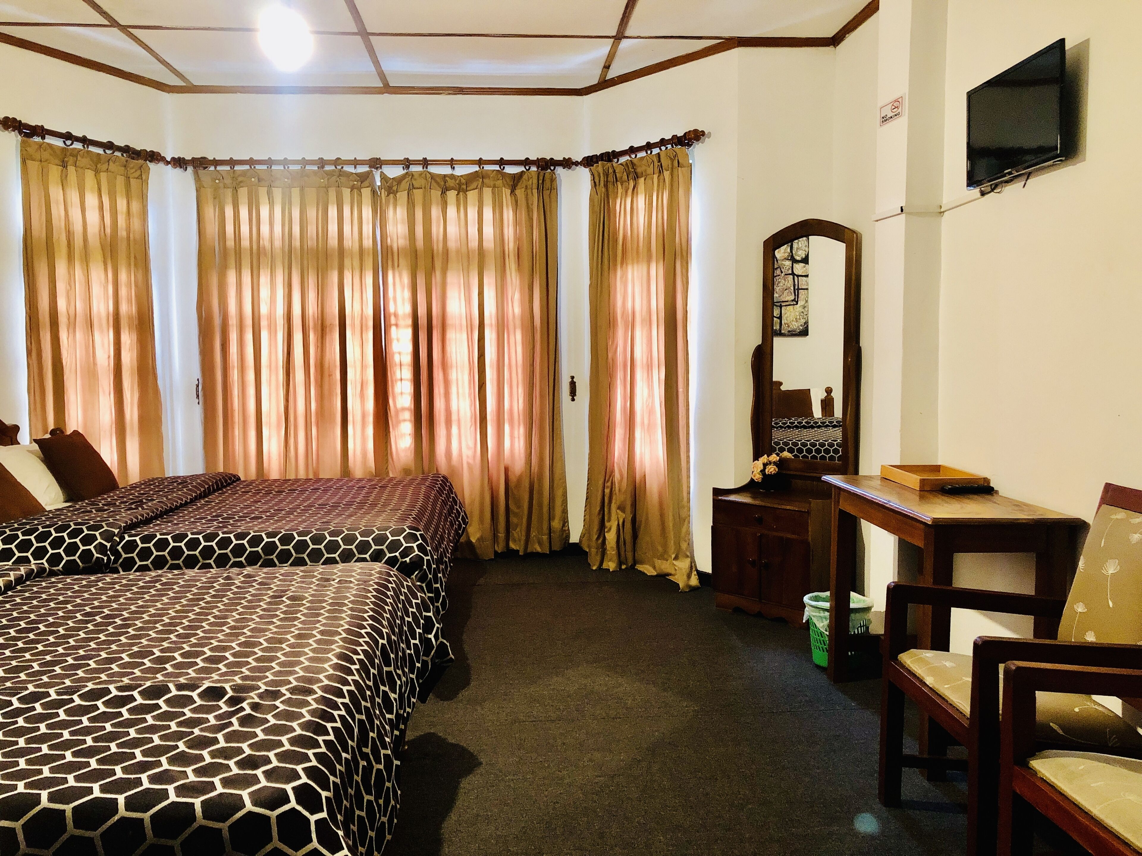 Quarto de hotel com duas camas, uma escrivaninha, uma cadeira, um espelho e cortinas.