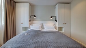 Quarto com cama, cabeceira branca, duas mesas de cabeceira e uma janela grande com cortinas.