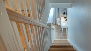 Uma escadaria moderna com degraus de madeira e corrimãos brancos, que leva a um quarto com uma escrivaninha, cadeira e luminária.