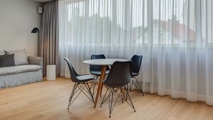 Uma sala de estar moderna com uma mesa redonda e quatro cadeiras, um sofá cinza e cortinas leves sobre uma grande janela.