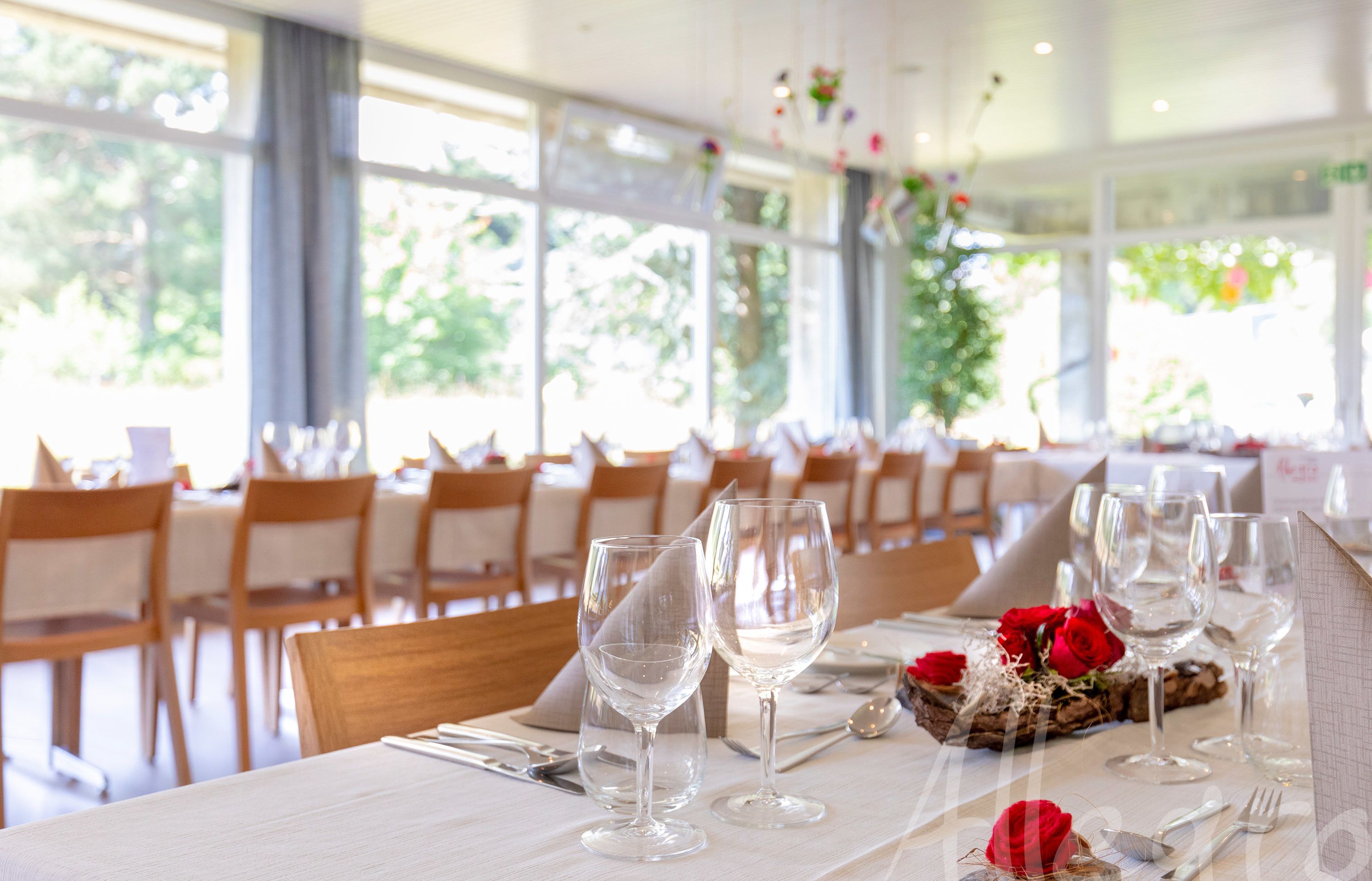 Restaurante com uma mesa comprida posta para uma refeição, com copos de vidro transparentes e um arranjo central de rosas vermelhas.
