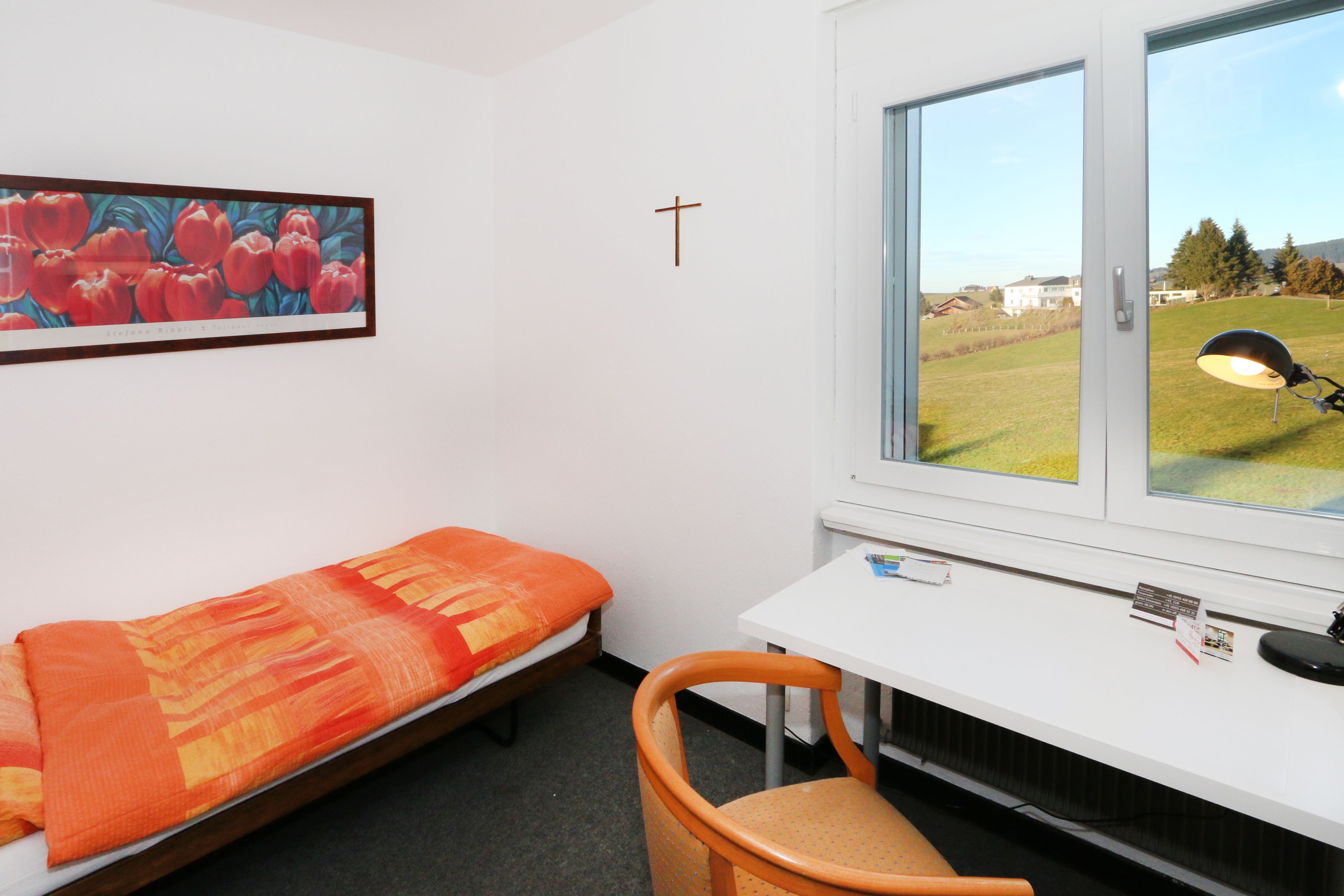 Quarto com cama, escrivaninha, cadeira, janela com vista para uma área gramada e uma cruz na parede.