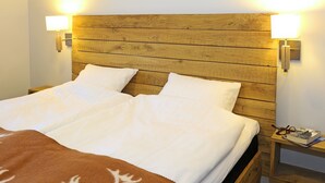 Uma cama com cabeceira de madeira, duas luminárias de apoio e uma mesinha de cabeceira com copos e um livro.