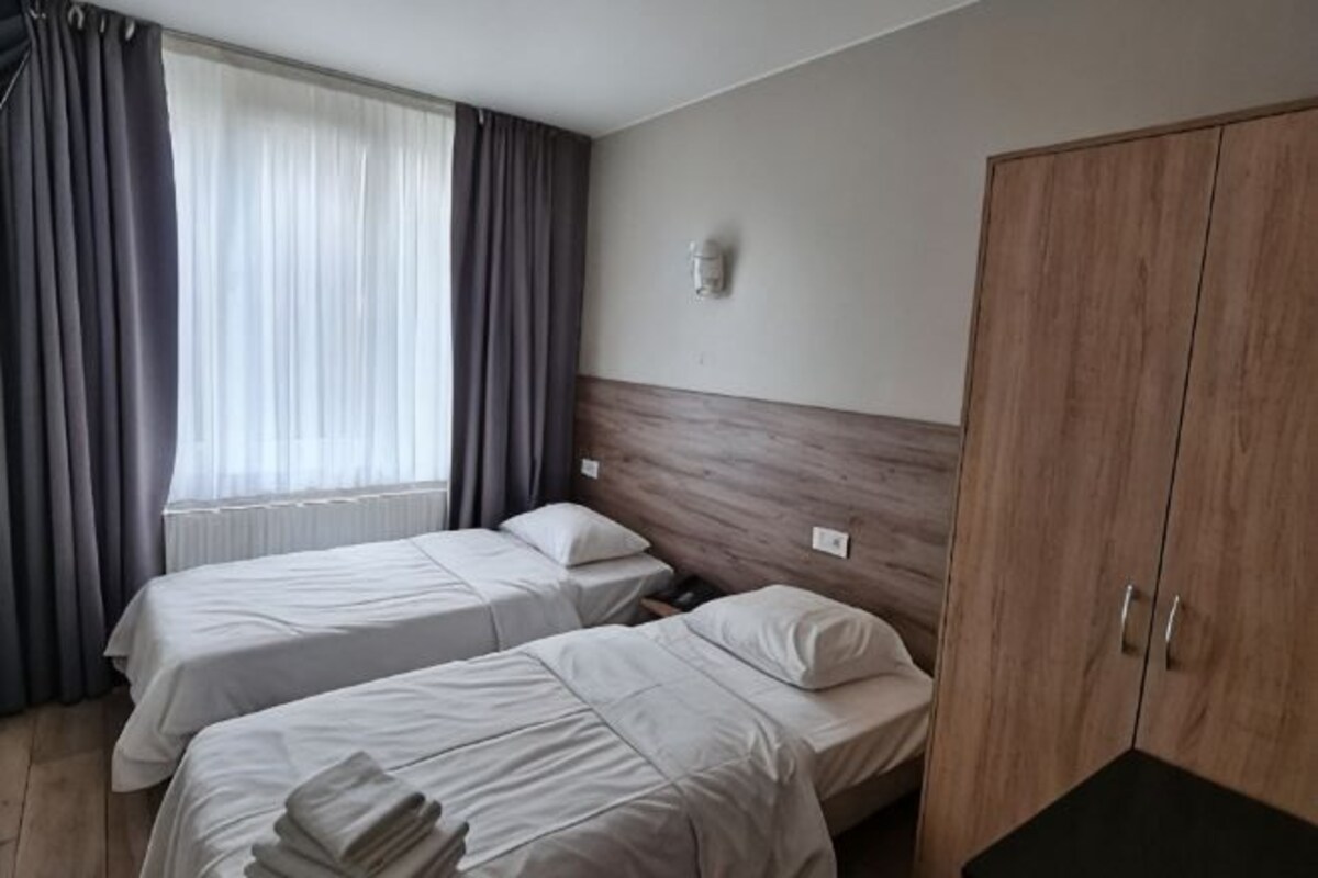 Quarto de hotel com duas camas, um guarda-roupa, uma janela com cortinas e uma luminária na parede.