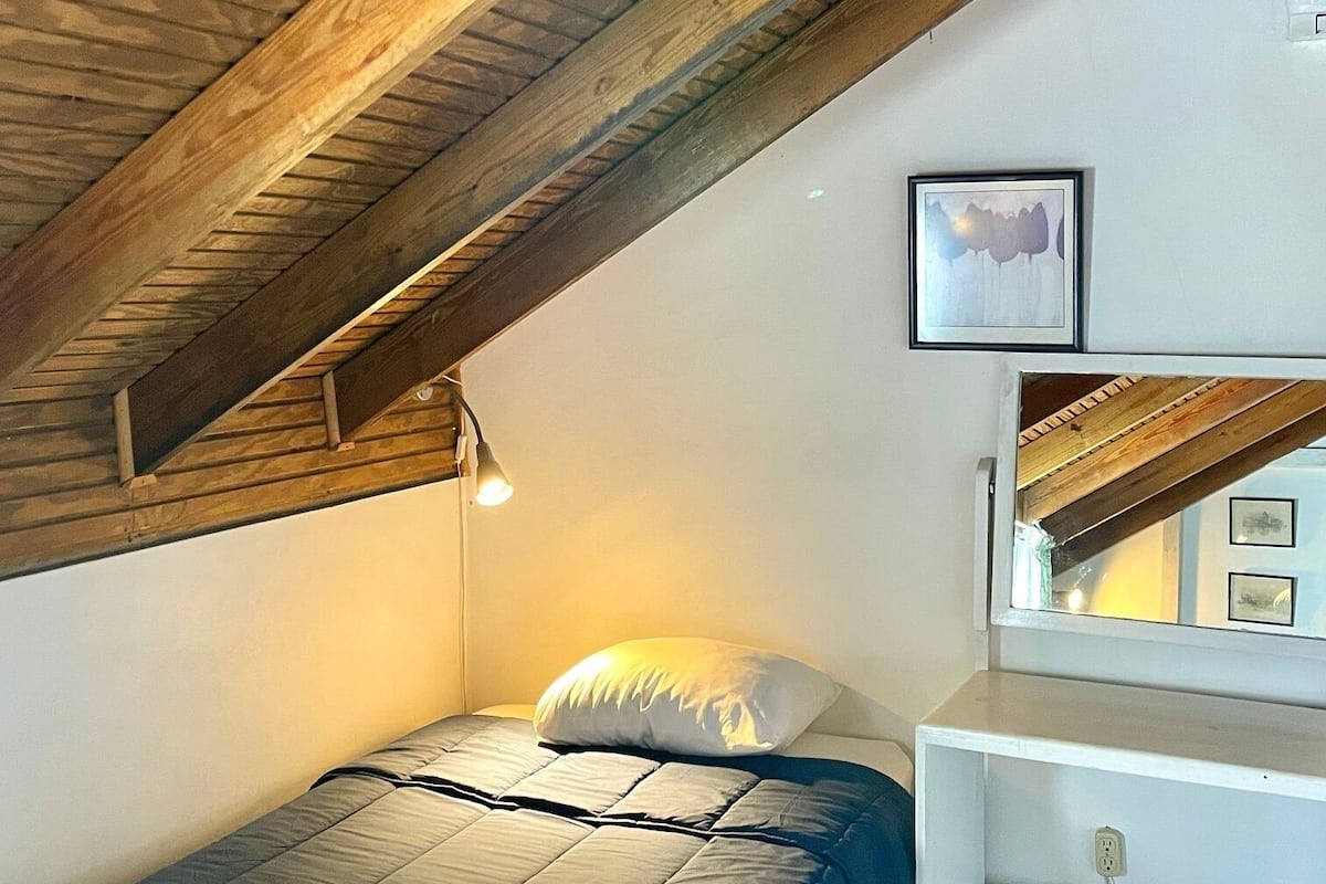 Quarto com teto inclinado, cama com colcha azul, cômoda branca e quadro na parede.