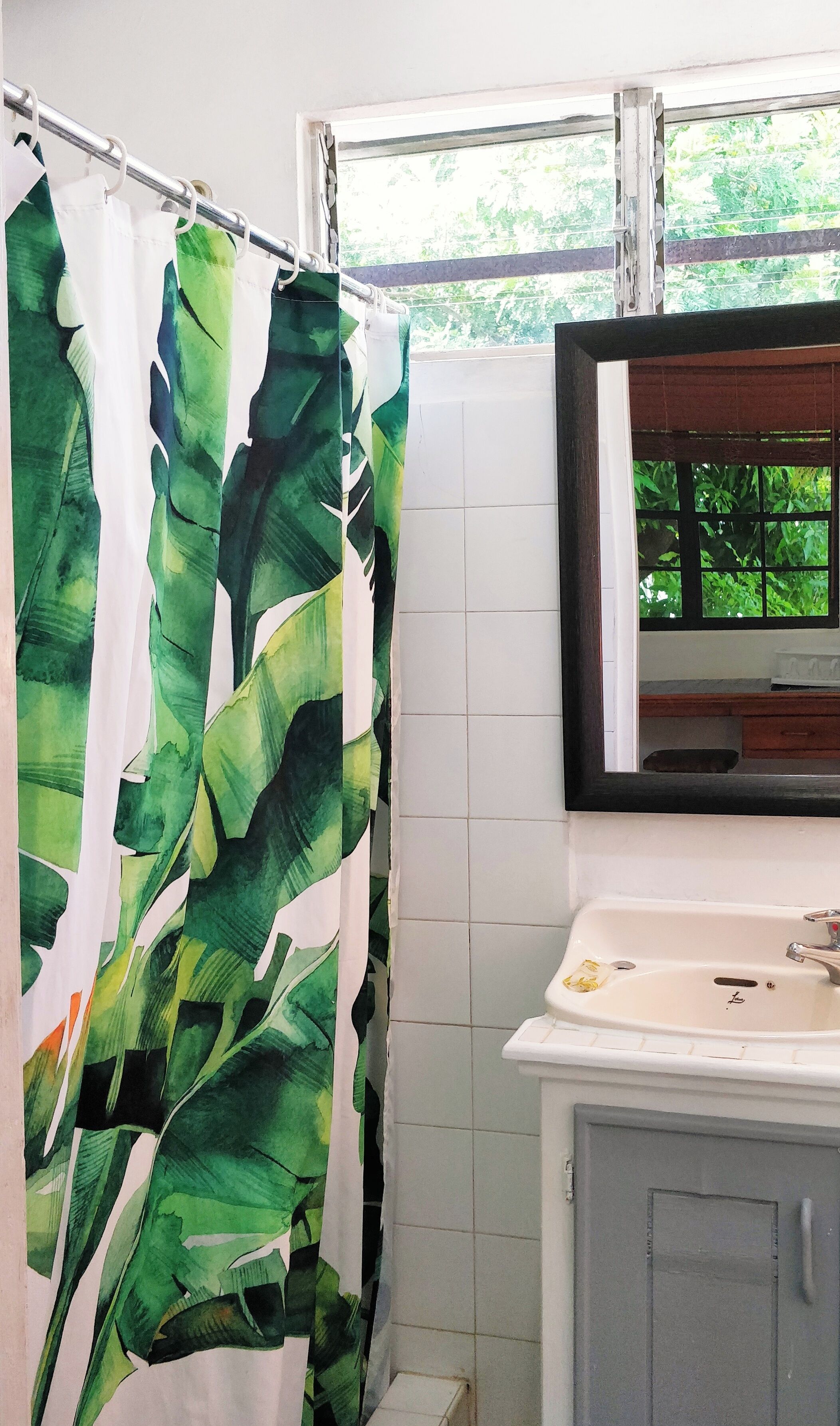 Casa de banho com cortina de chuveiro estampada com folhas verdes, pia branca e janela com vista para vegetação.