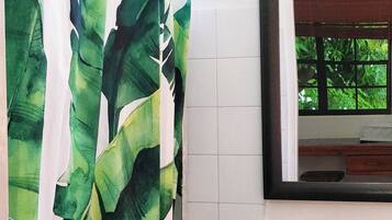 Casa de banho com cortina de chuveiro estampada com folhas verdes, pia branca e janela com vista para vegetação.