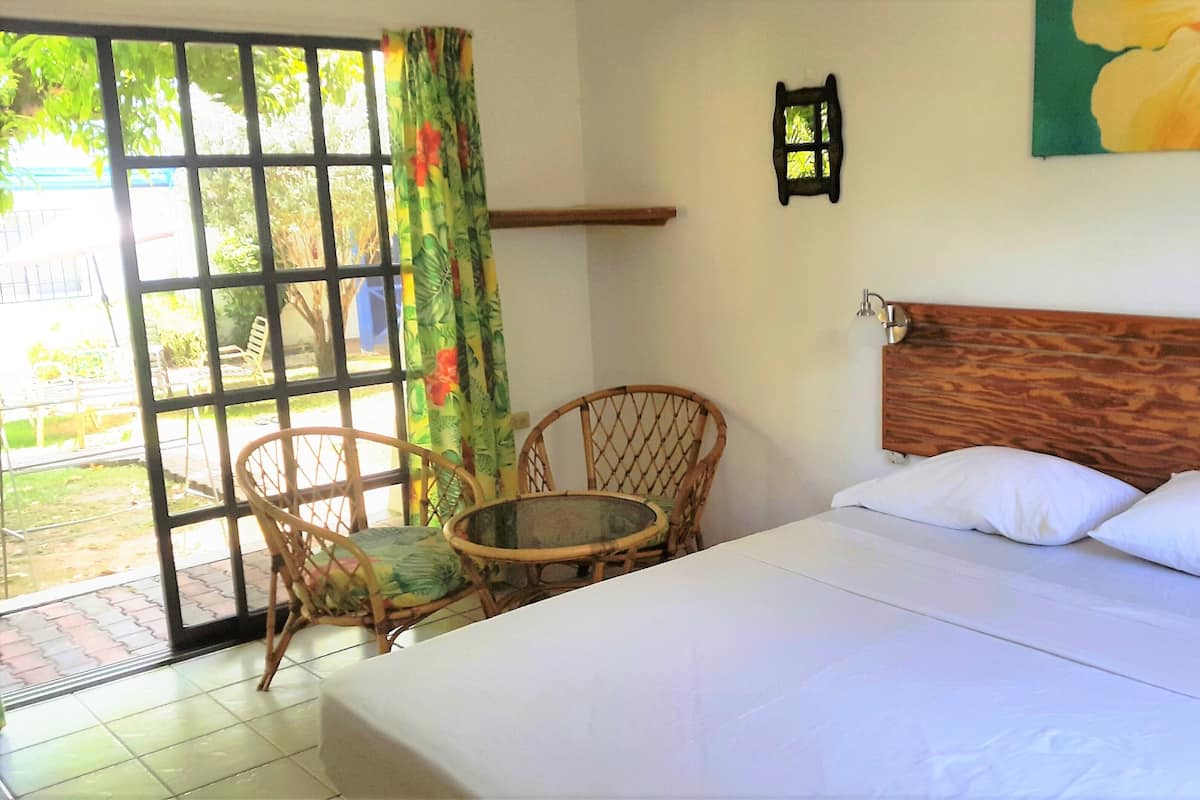 Quarto com cama, cadeira, mesa pequena, janela com cortinas e quadro na parede.