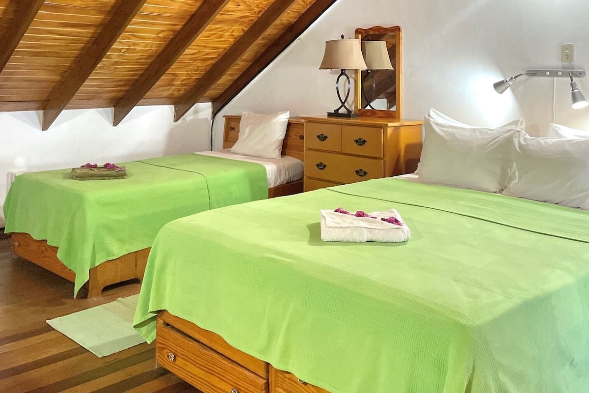 Quarto com duas camas, vigas de madeira no teto, mesa de cabeceira com abajur e criado-mudo.