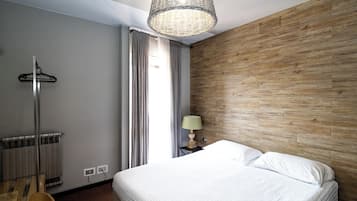Quarto de hotel com cama, parede de destaque em madeira, luminária pendente, radiador e janela com cortinas.