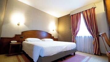 Quarto de hotel com cama, mesas de cabeceira, um criado-mudo, janela com cortinas e uma cadeira de madeira.