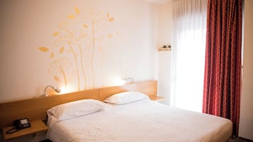 Quarto de hotel com cama, mesa de cabeceira, luminária na parede e janela com cortinas.