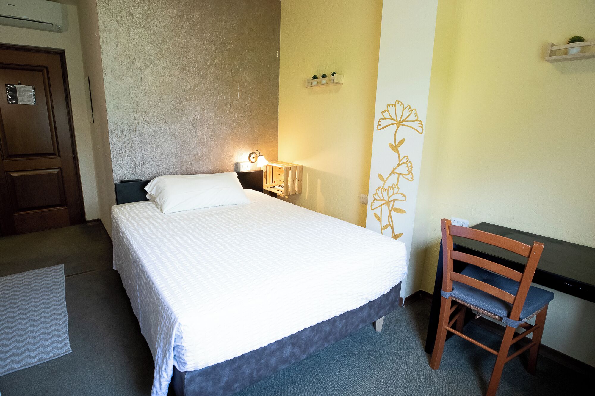 Quarto de hotel com uma cama, uma escrivaninha e uma cadeira. Há uma prateleira fixada na parede com plantas e uma peça decorativa.