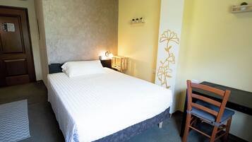 Quarto de hotel com uma cama, uma escrivaninha e uma cadeira. Há uma prateleira fixada na parede com plantas e uma peça decorativa.