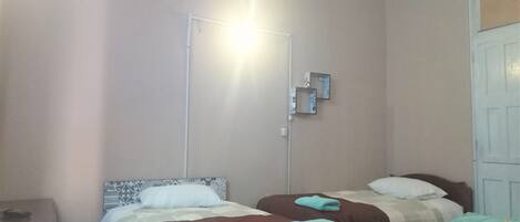 Quarto com duas camas, cada uma com um travesseiro e uma toalha dobrada. Há uma porta à direita e um interruptor de luz na parede.