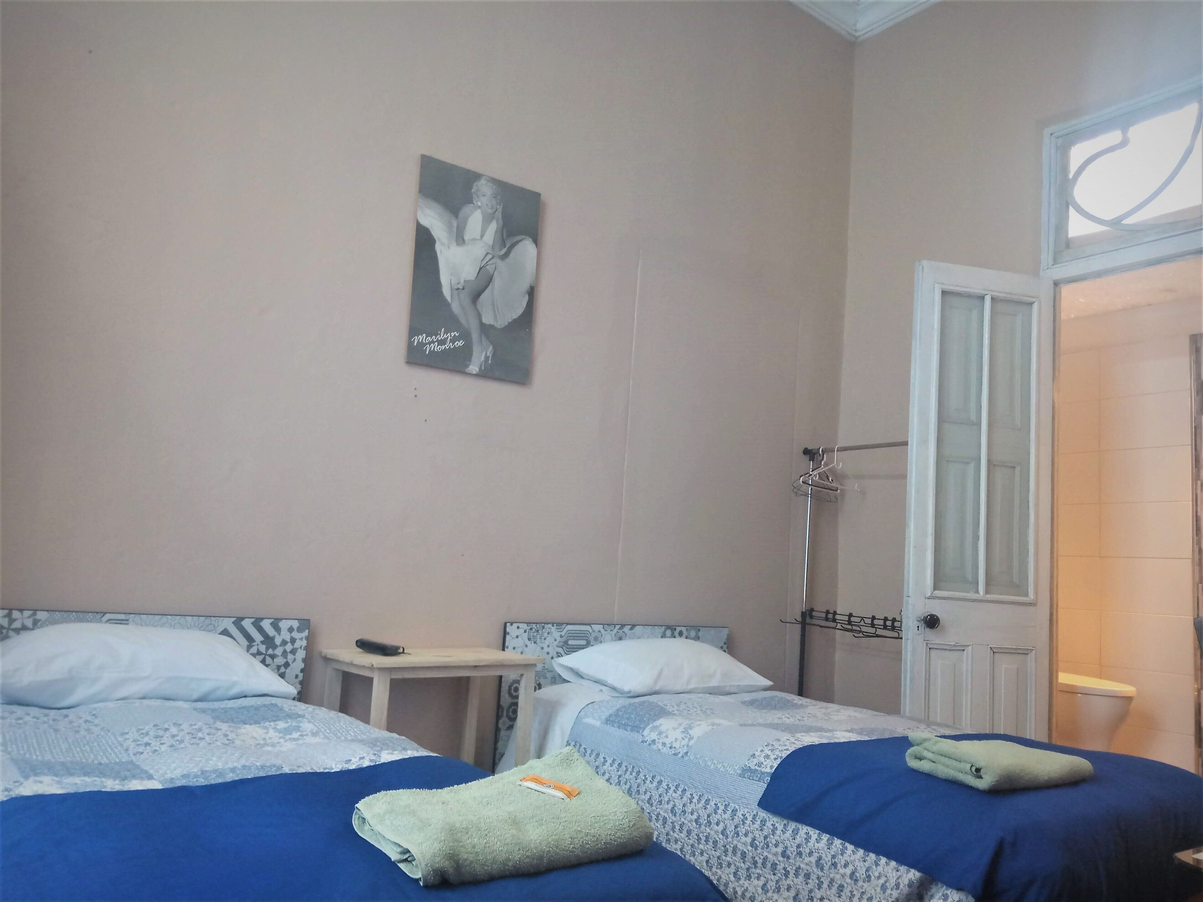 Quarto de hotel com duas camas, uma mesinha de cabeceira, área de chuveiro e uma porta.