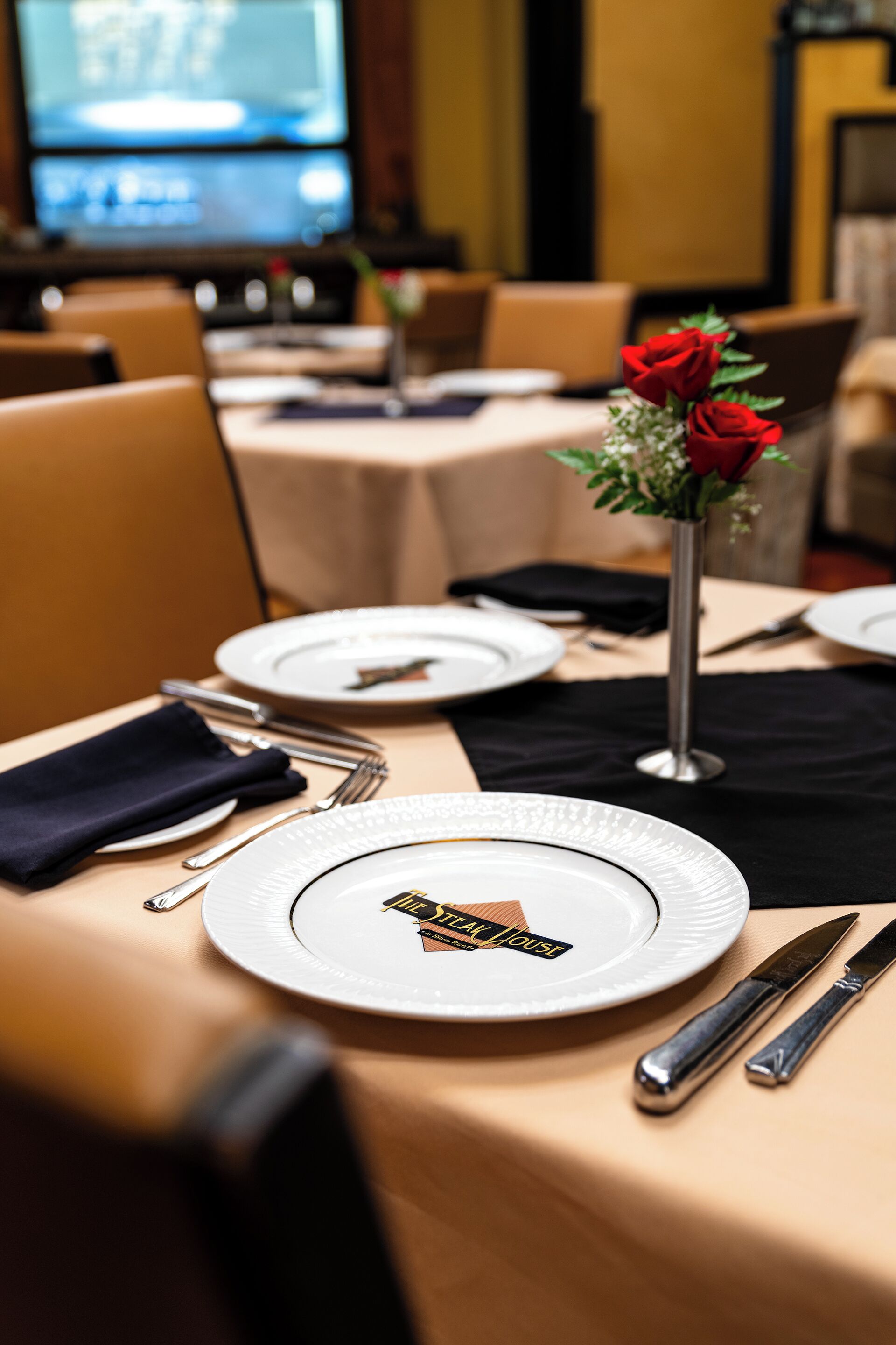 Uma mesa de jantar posta, com pratos, talheres e um vaso de rosas vermelhas.
