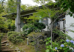 Uma casa tradicional japonesa com uma escadaria de madeira que leva à entrada, rodeada por vegetação exuberante e diversas plantas.