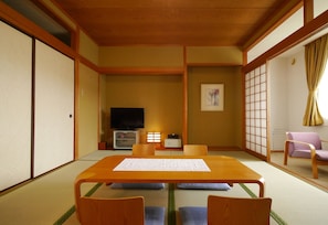Um quarto em estilo japonês tradicional com tatames, uma mesa de madeira baixa com almofadas, uma televisão e portas deslizantes.