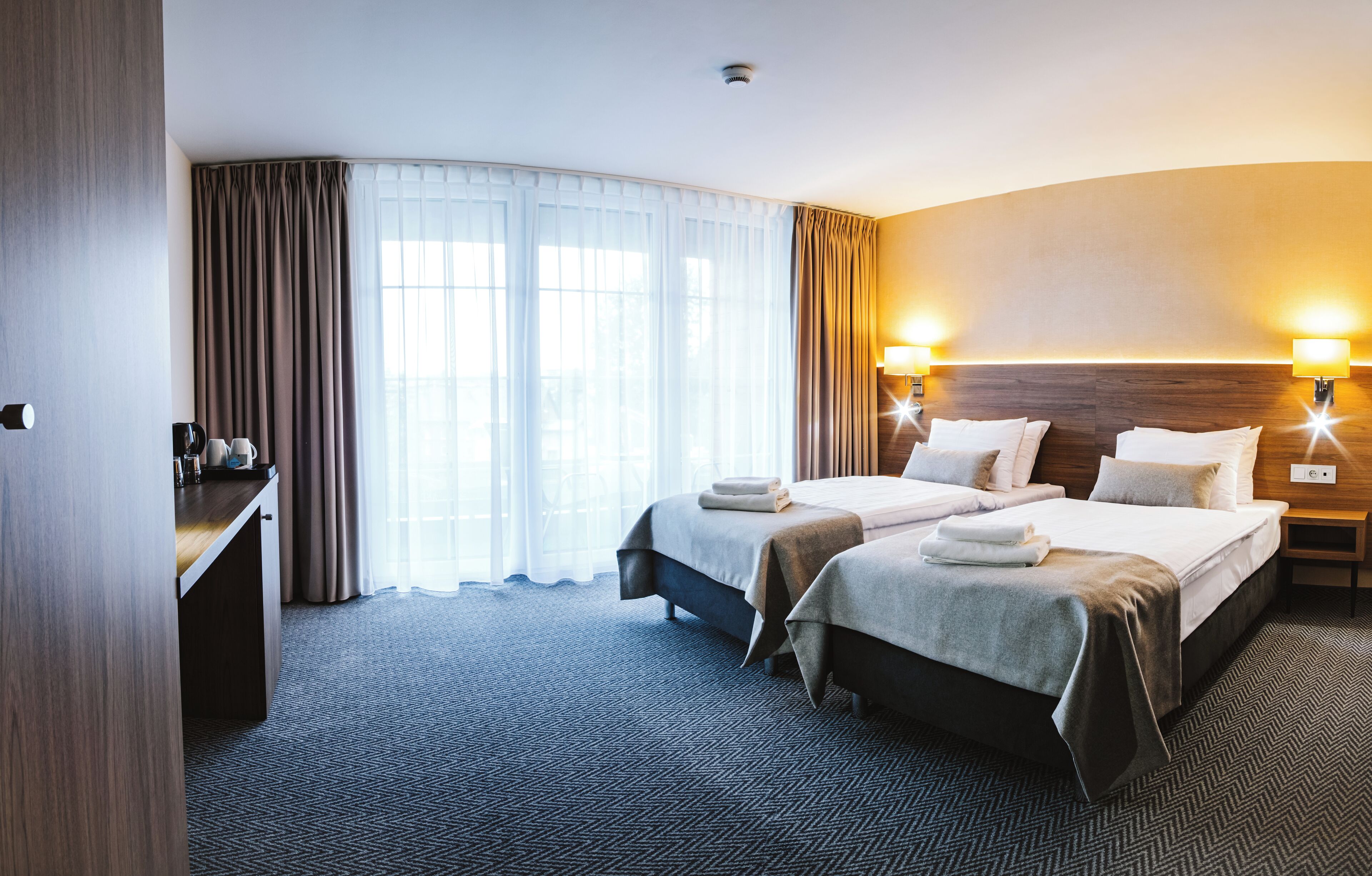 Quarto de hotel com duas camas, uma escrivaninha e uma janela grande com cortinas.