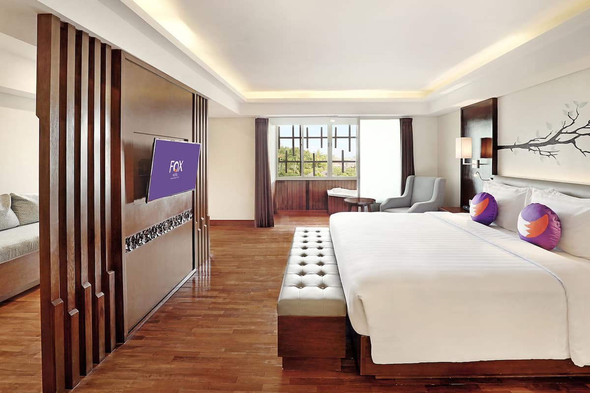 Quarto de hotel com uma cama grande, área de estar com mesa e televisão fixada em um painel de madeira.