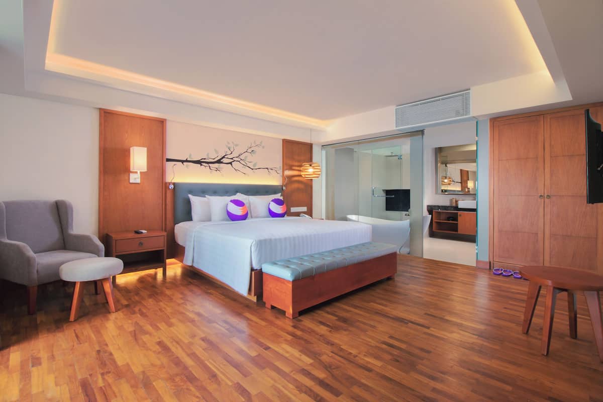 Quarto de hotel moderno com piso de madeira, cama com cabeceira, um banco, uma cadeira, guarda-roupa e banheiro visível por uma porta de vidro.