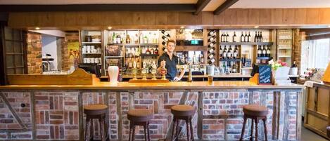 Uma área de bar com um bartender, balcão de madeira, banquetas e prateleiras com garrafas.