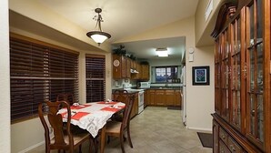 Uma sala de jantar com uma mesa posta para quatro pessoas, cadeiras de madeira, um lustre e uma cozinha ao fundo.