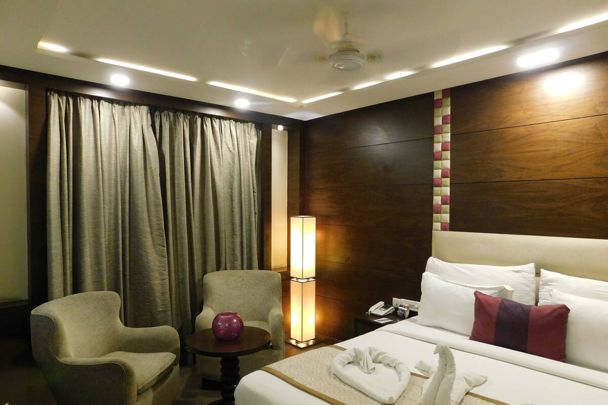 Quarto de hotel com cama, duas poltronas, mesa com um vaso, abajur e mesa de cabeceira com telefone e garrafa.