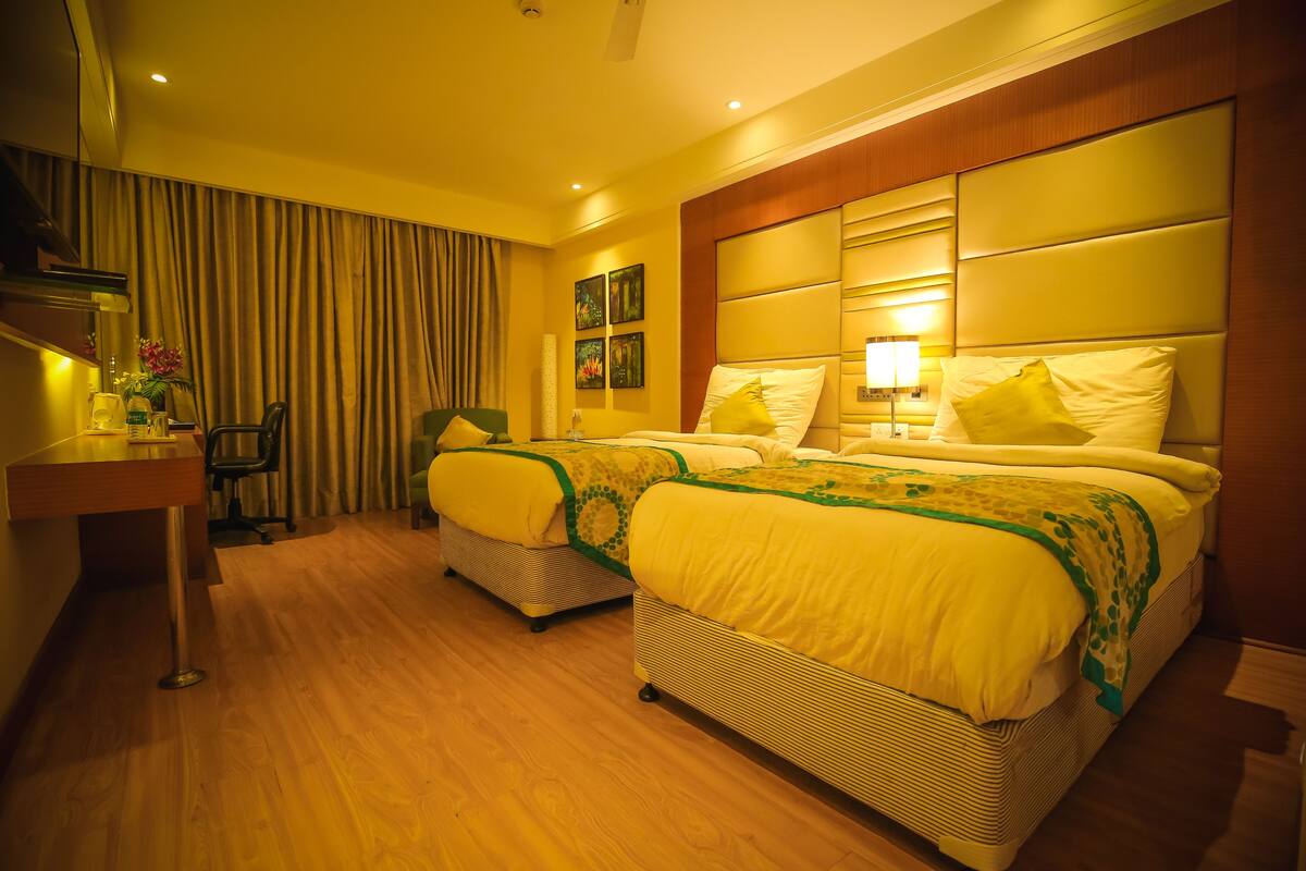 Quarto de hotel com duas camas, uma escrivaninha, uma cadeira e uma janela grande com cortinas.