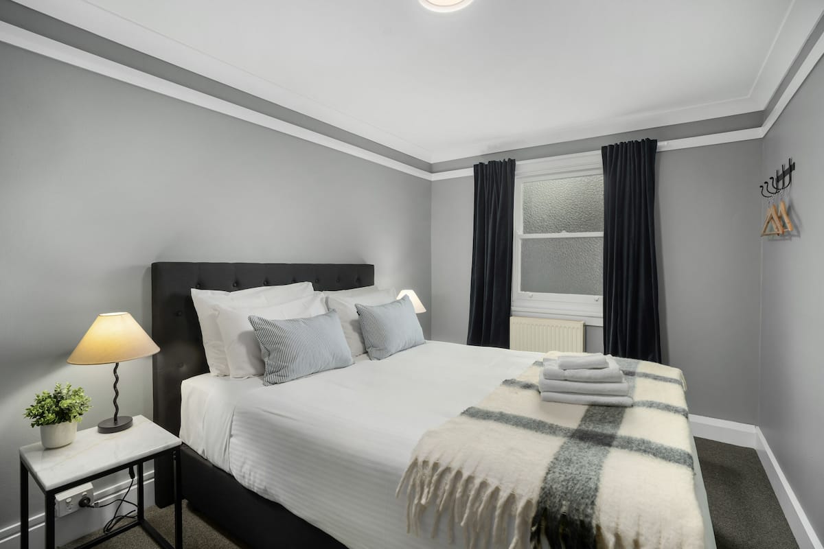 Quarto bem arrumado com cama, mesa de cabeceira, abajur e janela com cortinas.