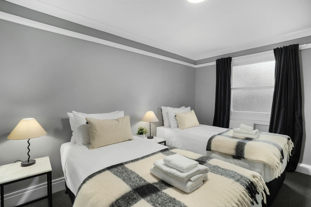Quarto de hotel com duas camas, janela com cortinas, luminária em cada lado e uma pequena planta no criado-mudo.