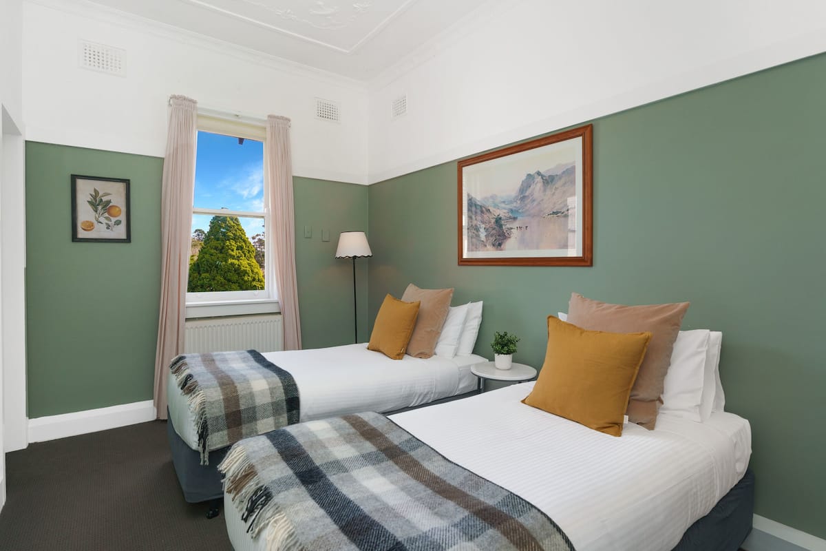 Quarto de hotel com duas camas, janela com cortinas, quadro na parede e uma mesinha com abajur.
