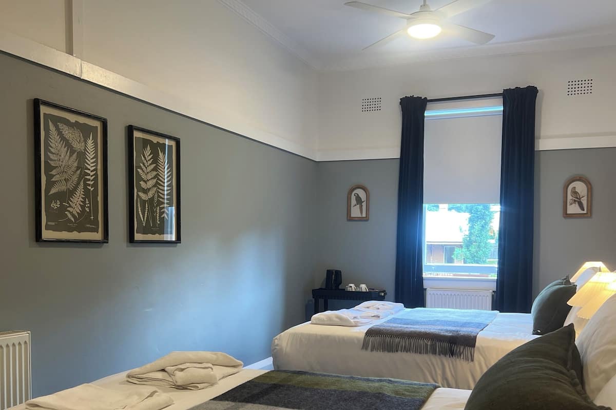 Quarto de hotel com duas camas, um ventilador de teto, impressões botânicas emolduradas na parede e uma janela com cortinas.