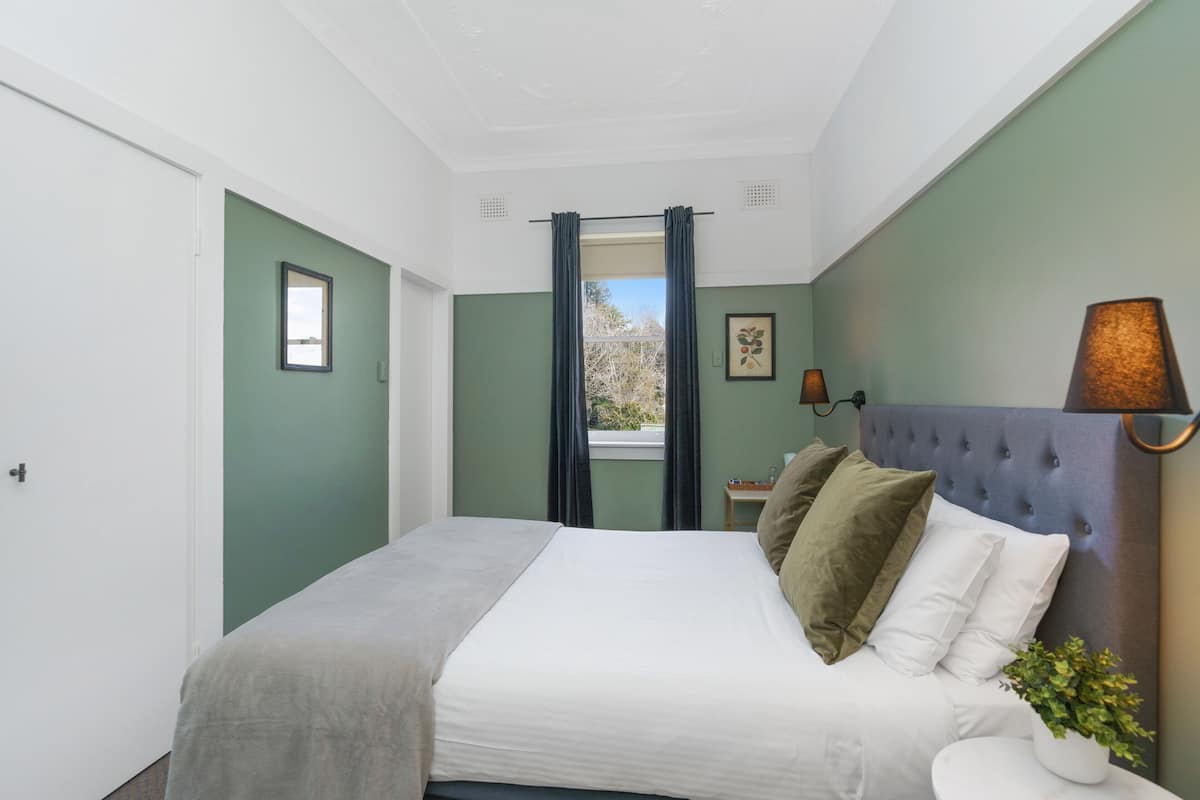 Quarto com cama, janela com cortinas, mesa de cabeceira e luminária na parede.