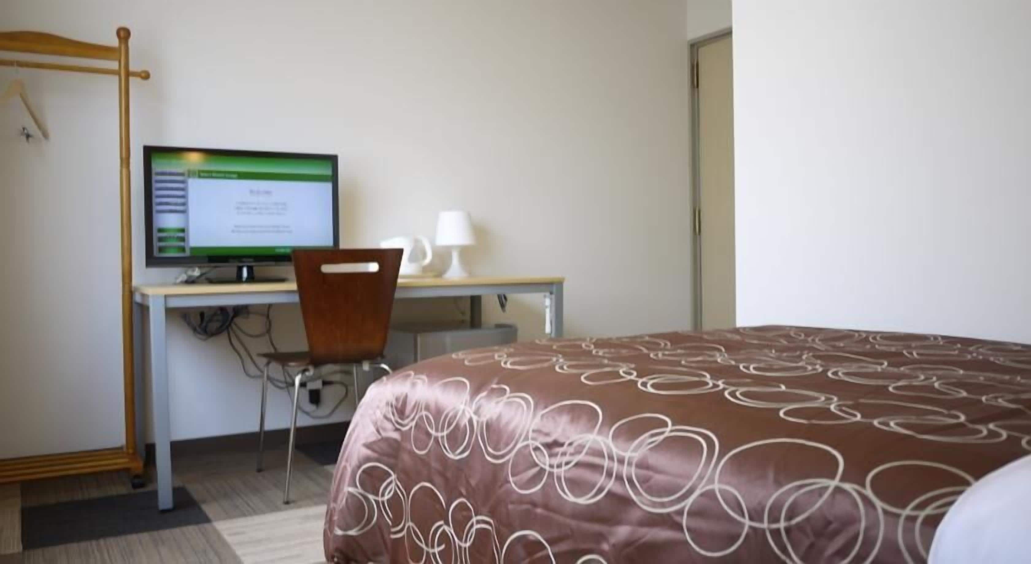 Quarto de hotel com cama, escrivaninha com computador, cadeira e luminária.