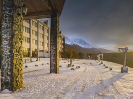 Área externa coberta de neve, com pilares de pedra, um edifício com grandes janelas e uma montanha ao fundo.