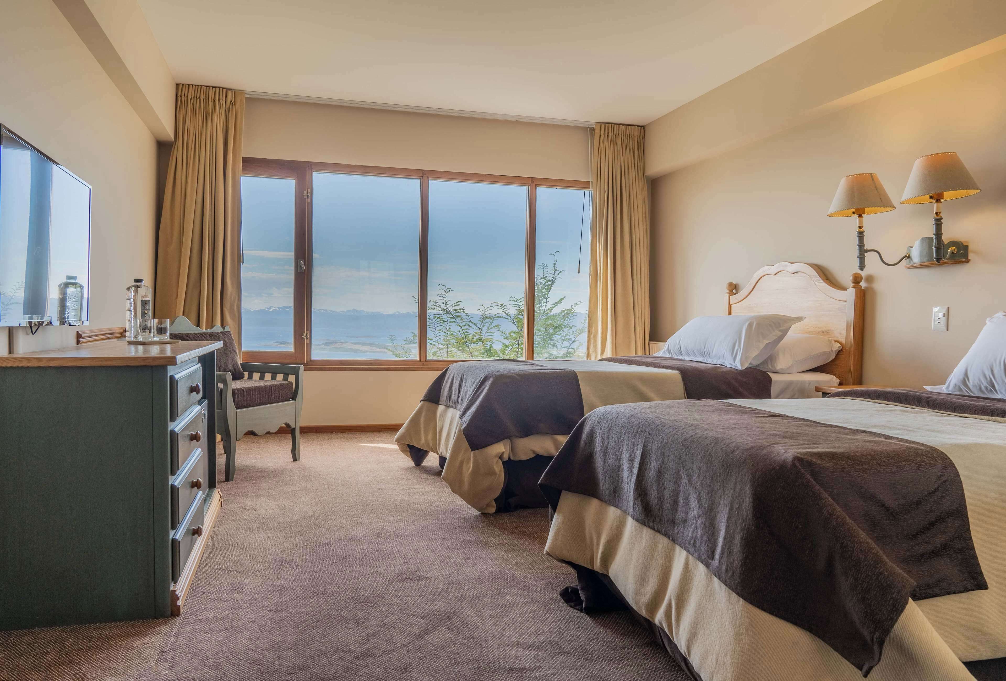 Quarto de hotel com duas camas, uma escrivaninha, um armário e uma janela grande com vista para o exterior.