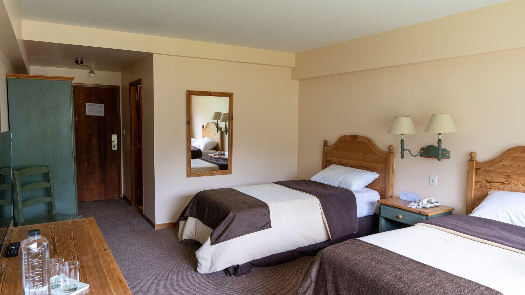 Quarto de hotel com duas camas, uma escrivaninha com abajur, um espelho e um guarda-roupa.
